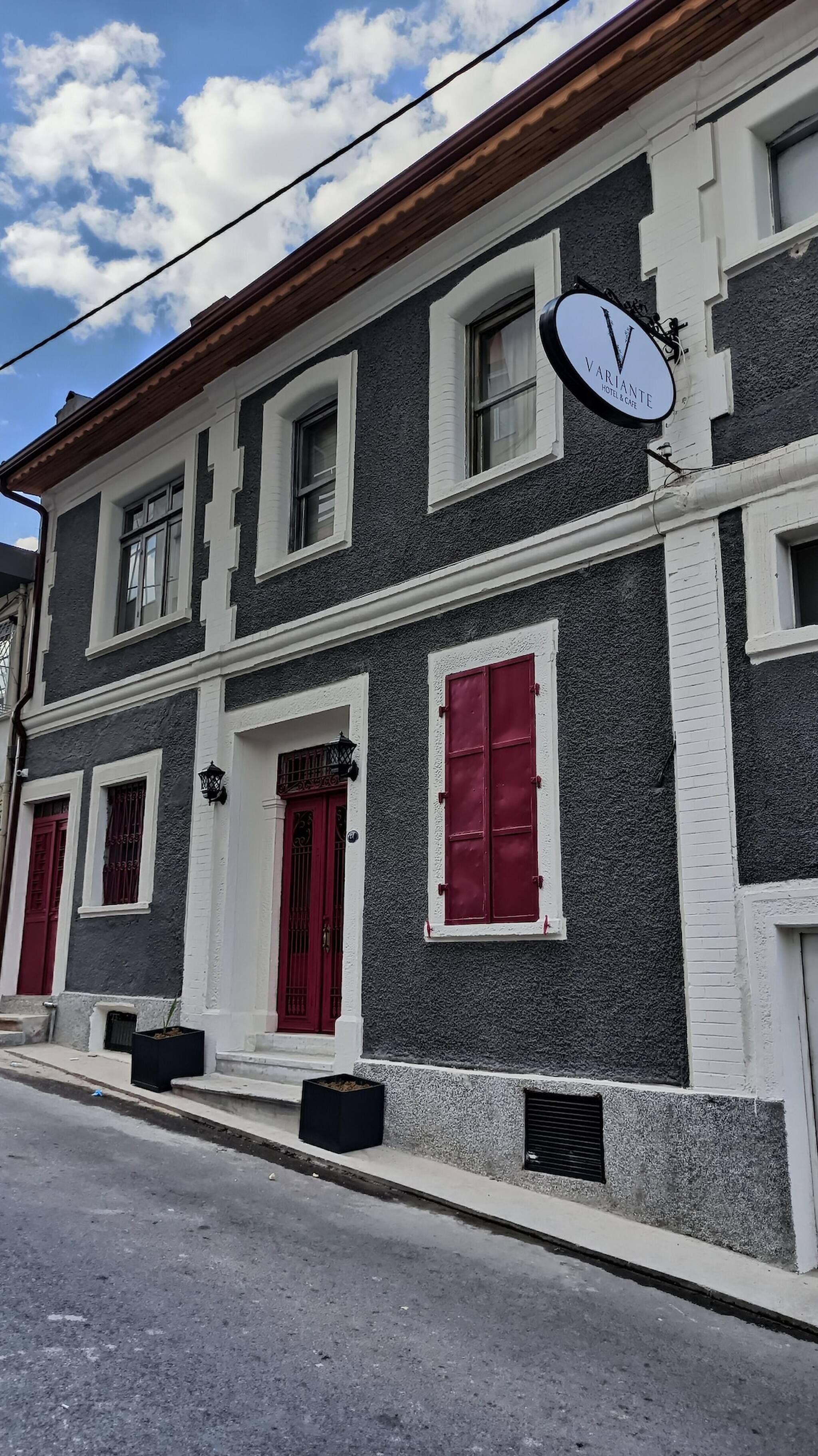 Um edifício de dois andares com uma placa indicando hotel e café. Várias janelas com persianas, uma porta visível e uma pequena área externa com vasos de plantas.