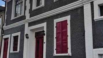 Um edifício de dois andares com uma placa indicando hotel e café. Várias janelas com persianas, uma porta visível e uma pequena área externa com vasos de plantas.