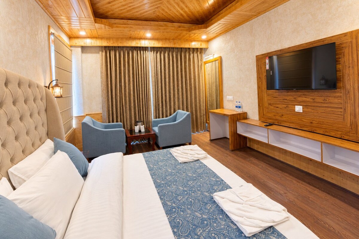 Quarto de hotel com uma cama grande, duas poltronas, uma televisão e teto de madeira.