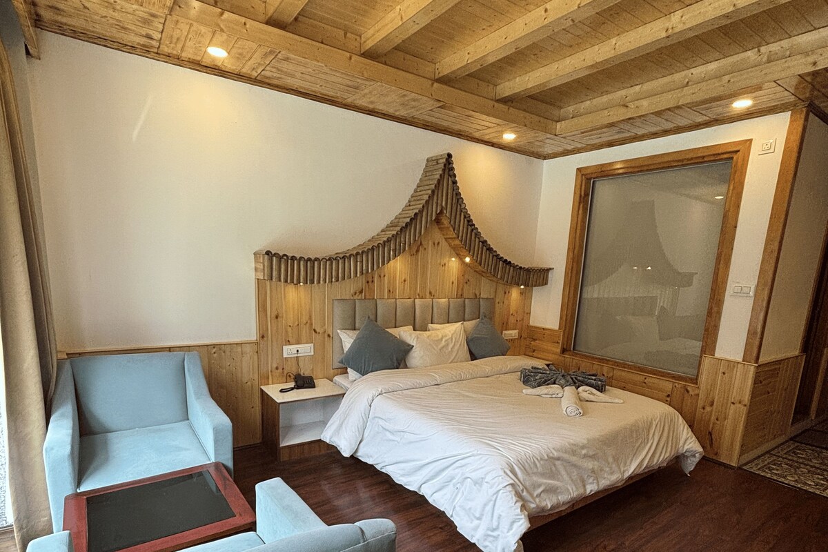 Quarto com teto de madeira, cama com almofadas, mesa de cabeceira, cadeira e janela ampla.