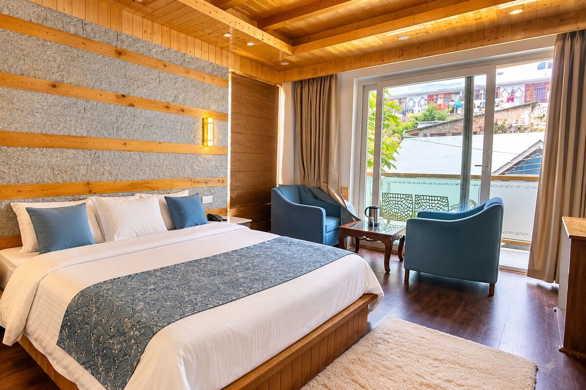 Quarto de hotel com cama grande, cabeceira de madeira, área de estar com uma cadeira e mesa, e vista para o exterior.