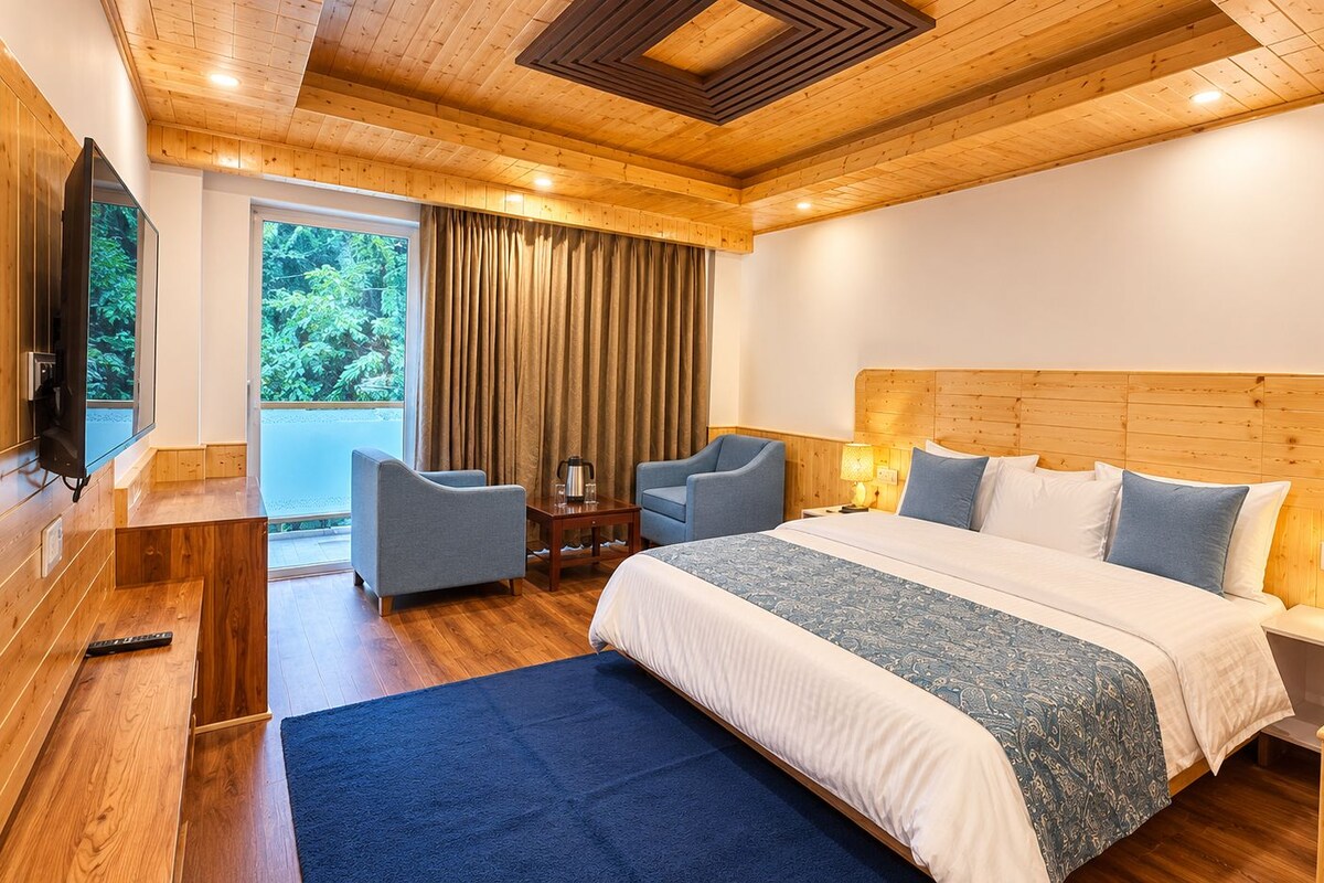 Quarto de hotel com uma cama grande, duas poltronas, uma TV e vista para o verde lá fora.