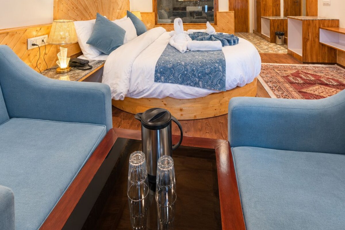 Quarto de hotel com teto de madeira, cama com roupa de cama azul, duas poltronas, mesa de centro com copos e banheiro visível através de uma porta de vidro.