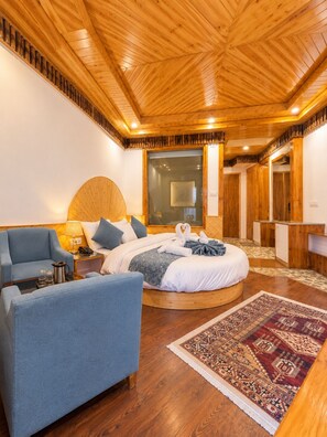 Quarto de hotel com teto de madeira, cama redonda, poltrona azul, mesa de cabeceira com abajur e um espelho grande.