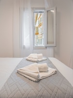 Quarto de hotel com cama, duas toalhas, janela com cortinas e vista para edifícios.