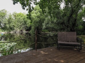 Um deck de madeira com um banco, cercado por uma barreira de corda, com vista para um corpo d'água calmo com vitórias-régias e vegetação densa.