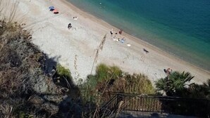 Uma praia costeira com águas cristalinas, areal arenoso e montanhas ao fundo.