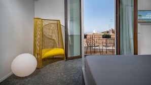 Quarto de hotel com cama, uma cadeira amarela, um objeto branco arredondado e uma varanda com móveis de exterior.