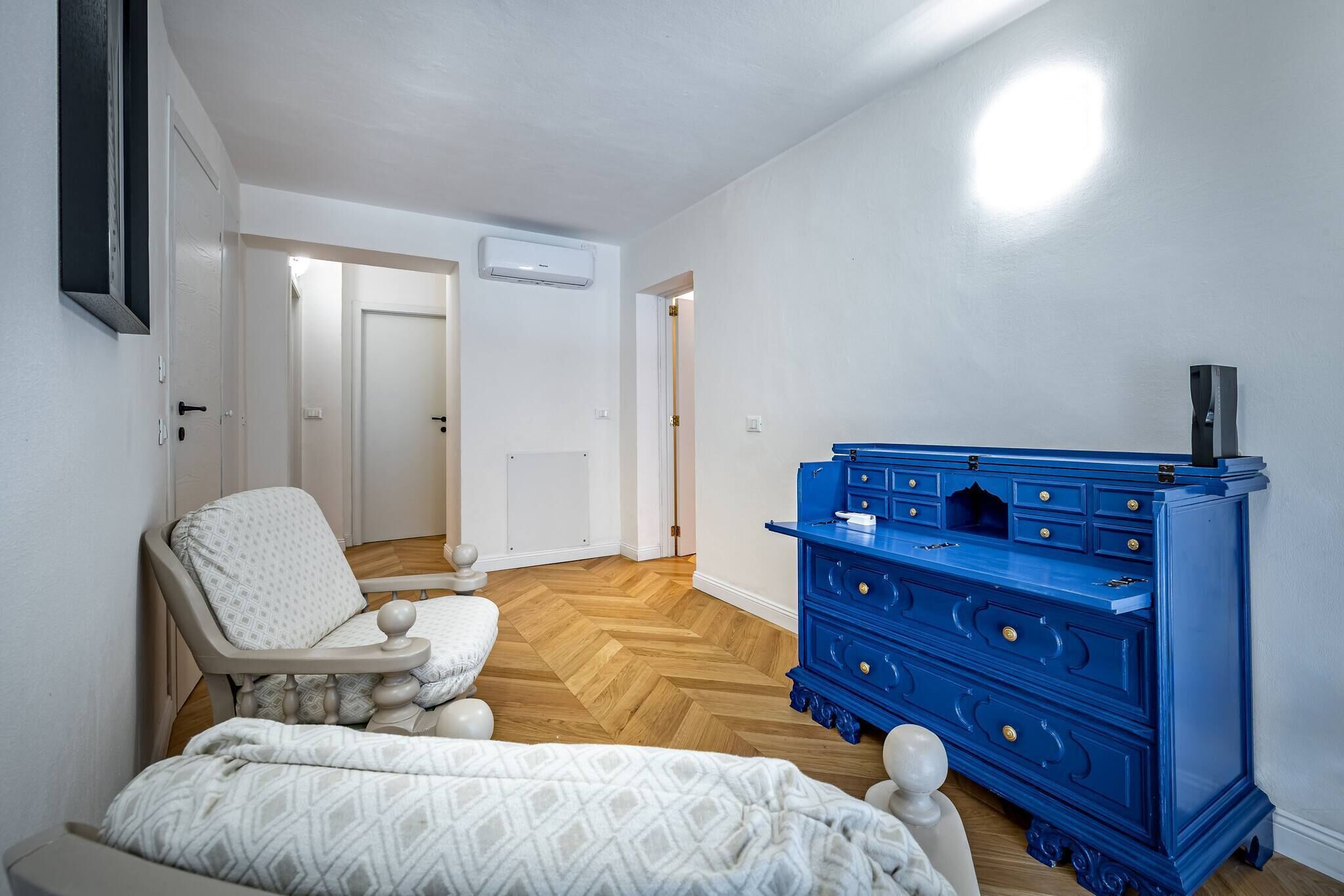 Quarto com soalho de madeira em padrão herringbone, cómoda azul, cadeira branca e cama com roupa de cama estampada.