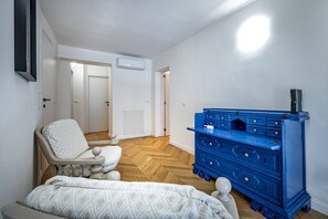 Quarto com soalho de madeira em padrão herringbone, cómoda azul, cadeira branca e cama com roupa de cama estampada.