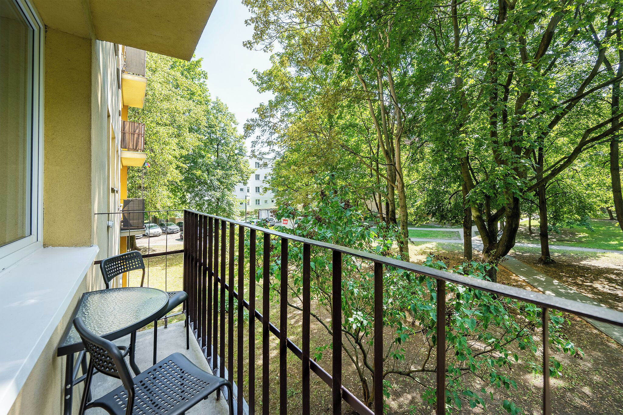 Uma varanda com mesa e cadeiras, com vista para um parque com árvores e um edifício ao fundo.