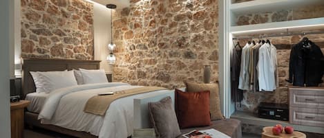 Quarto com parede de pedra, cama com roupa de cama branca, mesa de cabeceira com abajur e armário com roupas penduradas.
