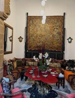 Uma sala de estar tradicional com um grande tapeçário, sofás ricamente decorados, uma mesa redonda vermelha e um espelho ornamental.