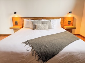 Cama bem arrumada com edredom branco, manta cinza e várias almofadas estampadas. Há duas mesas de cabeceira com luminárias e uma cabeceira de madeira.