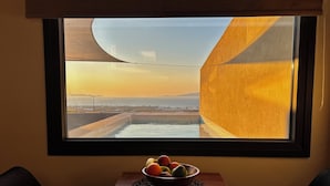 Um quarto com uma mesa e uma tigela de frutas, uma janela com vista para a paisagem e parte de uma cadeira.