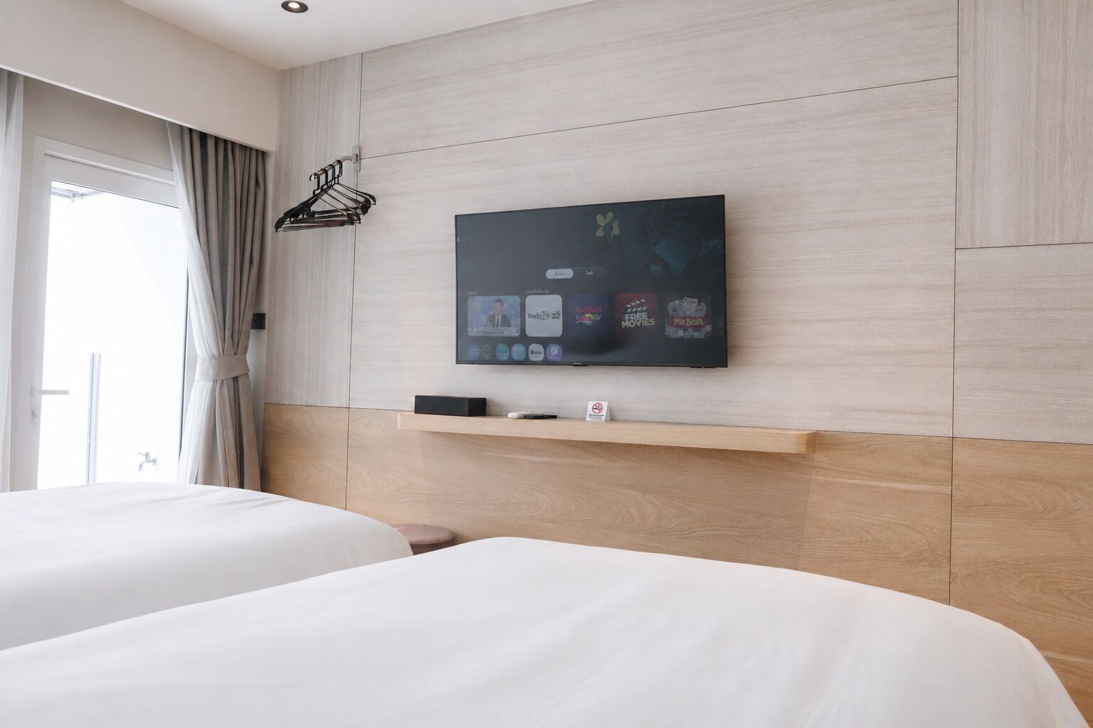 Quarto de hotel com duas camas, uma televisão fixada na parede exibindo diversos canais e uma estante de madeira abaixo.