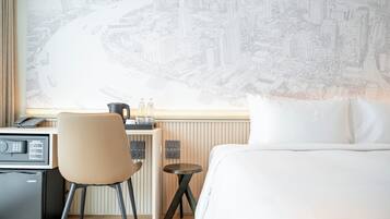 Quarto de hotel com cama, escrivaninha com cadeira, mesa pequena, chaleira e uma pintura de paisagem urbana na parede.