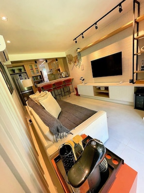Uma sala de estar moderna com sofá, televisão e área de cozinha ao fundo.