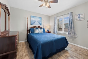 Quarto com cama de madeira, ventoinha no teto, pintura de um cais e janela com cortinas estampadas.