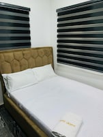 Quarto de hotel com uma cama, cabeceira e duas janelas com persianas.