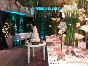 Área de recepção de casamento com um bolo de noiva em vários níveis, arranjos florais e uma variedade de sobremesas em uma mesa.