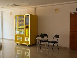 Uma máquina de vendas automática, uma pequena mesa com uma cadeira e um vaso de planta em um espaço interno limpo e bem iluminado.