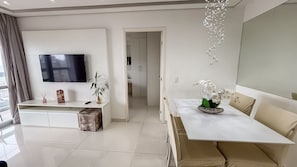 Uma sala de estar moderna com uma televisão de tela plana, uma mesa de jantar com um vaso de orquídeas brancas e um sofá.