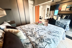 Quarto de hotel moderno com uma cama grande, uma cama menor, uma escrivaninha e uma cadeira. Há uma janela com vista para a praia.