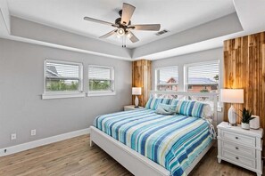 Quarto com cama, ventoinha no teto, duas janelas com persianas, mesa de cabeceira e parede com painéis de madeira vertical.