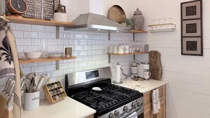 Uma cozinha com painel de azulejos brancos, eletrodomésticos de aço inoxidável, prateleiras de madeira e objetos decorativos.