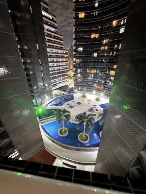 Um edifício alto com vários andares, um pátio central com palmeiras e um caminho iluminado com luzes verdes.