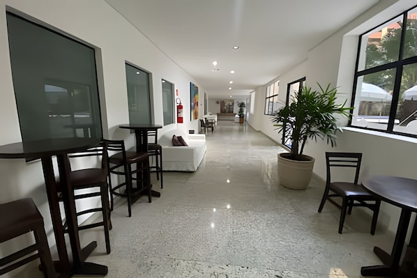 Um corredor espaçoso com um sofá branco, uma mesa de jantar e cadeiras, uma grande planta em vaso e um extintor de incêndio na parede.