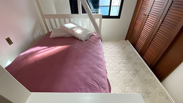 Quarto pequeno com uma cama de solteiro, cabeceira branca, colcha rosa e janela com persianas.