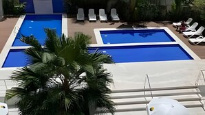 Uma área de piscina com duas piscinas retangulares, cadeiras de descanso e uma palmeira.