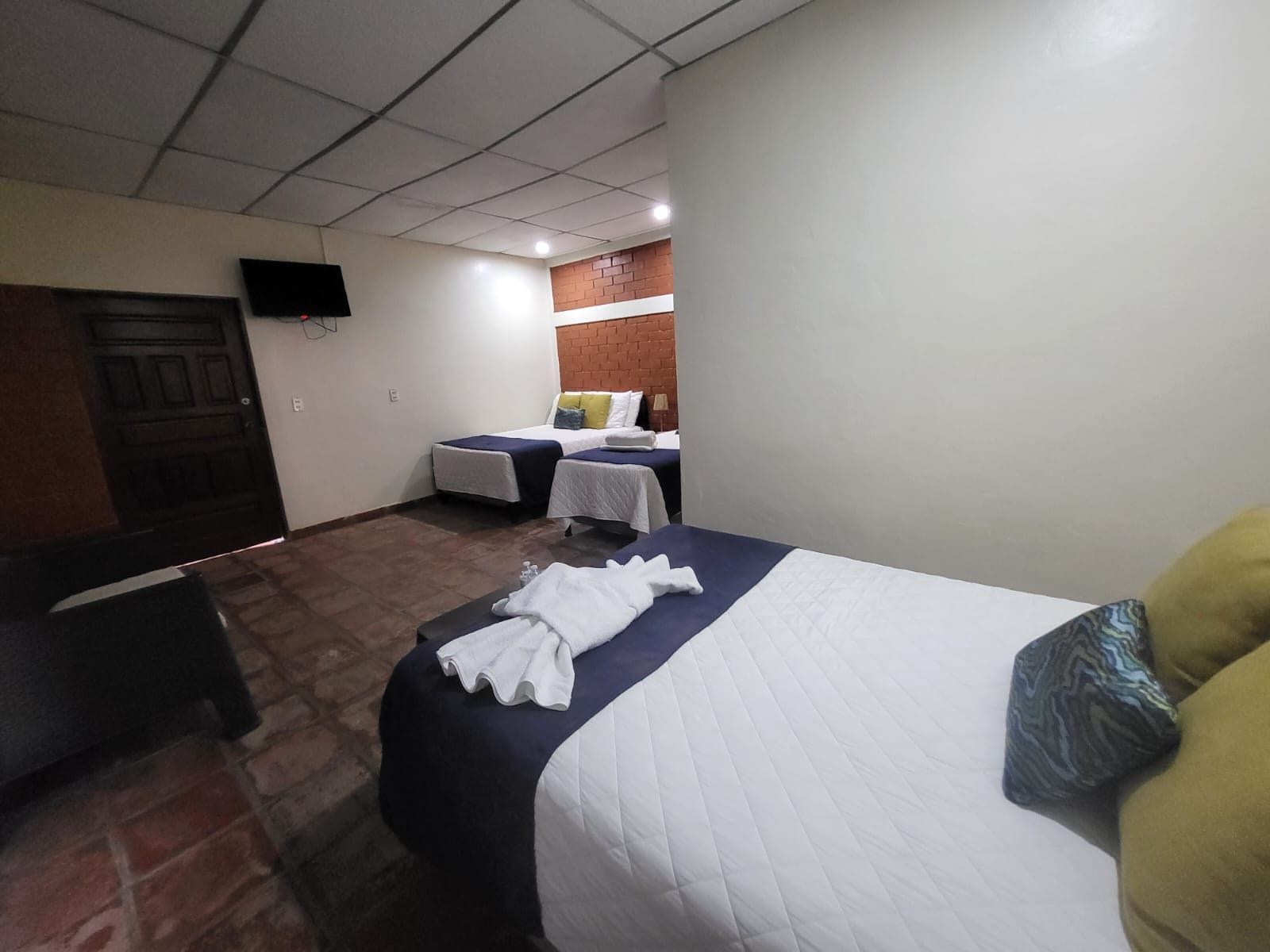 Quarto de hotel com duas camas, televisão na parede e parede decorativa de tijolos.