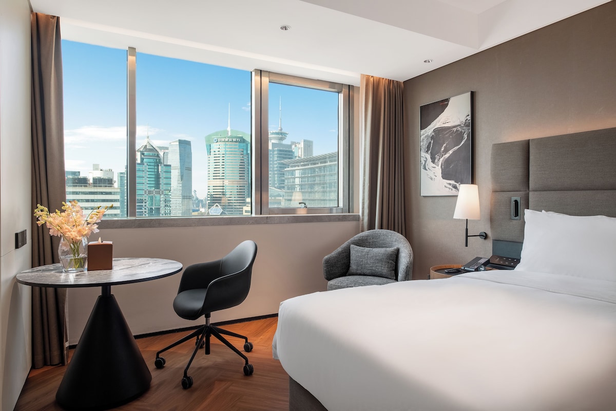 Quarto de hotel com cama, escrivaninha, cadeira, abajur e vista para a cidade através de amplas janelas.