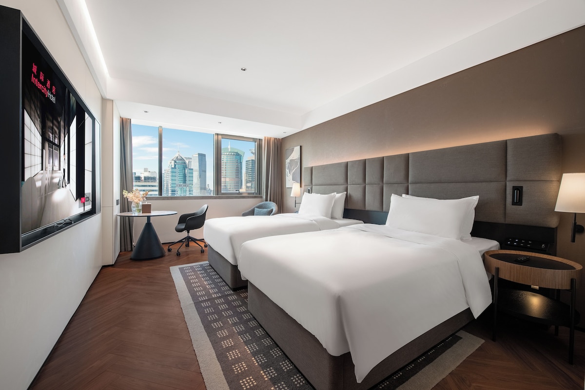 Quarto de hotel com duas camas, uma escrivaninha, uma cadeira, uma televisão e vista para a paisagem urbana.