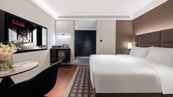 Quarto de hotel com cama, escrivaninha, cadeira, uma pequena mesa com um vaso de flores e televisão na parede.