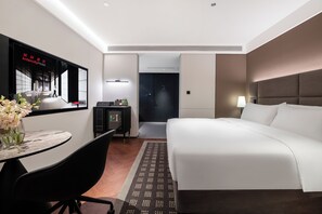 Quarto de hotel com cama, escrivaninha, cadeira, uma pequena mesa com um vaso de flores e televisão na parede.