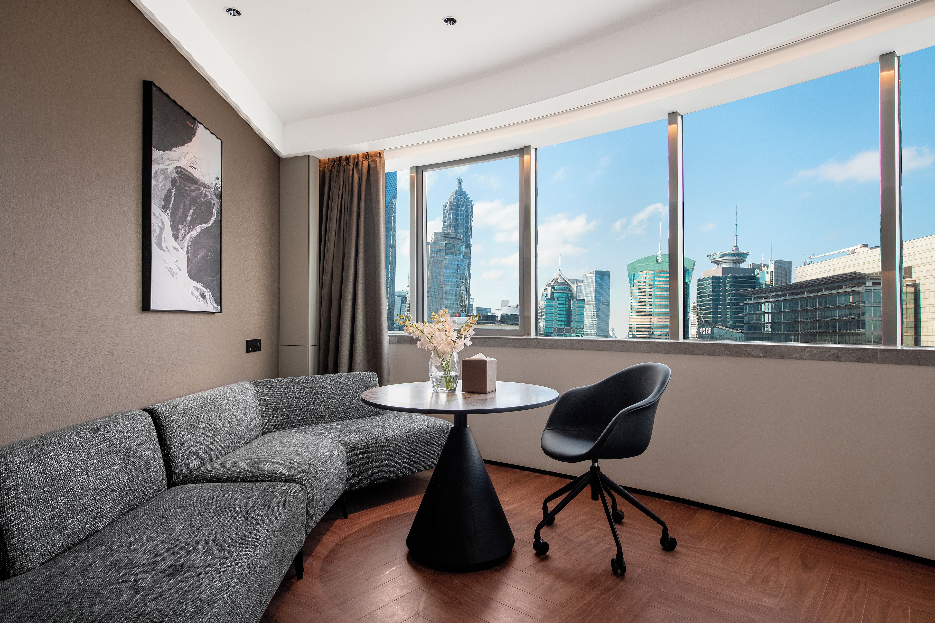 Quarto moderno com sofá seccional cinza, uma mesa redonda com um vaso de flores e vista para o horizonte urbano através de grandes janelas.