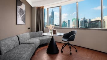 Quarto moderno com sofá seccional cinza, uma mesa redonda com um vaso de flores e vista para o horizonte urbano através de grandes janelas.
