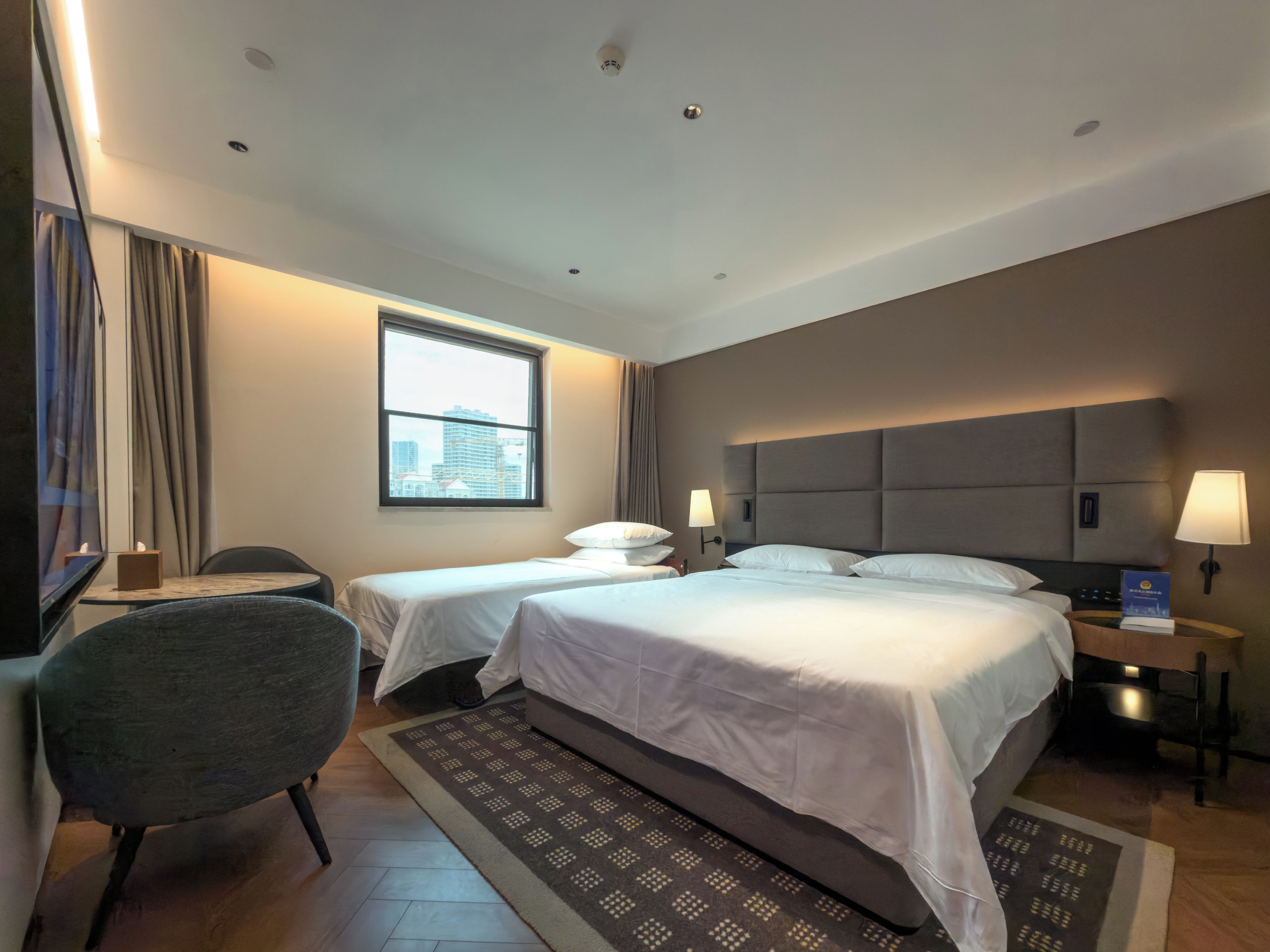 Quarto de hotel com duas camas, uma escrivaninha, uma cadeira, janela com vista e abajur na mesa de cabeceira.