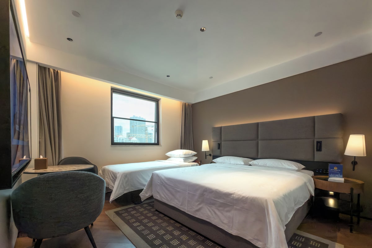 Quarto de hotel com duas camas, uma escrivaninha, uma cadeira, janela com vista e abajur na mesa de cabeceira.
