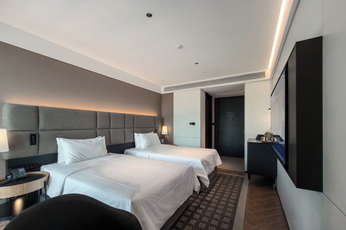 Quarto de hotel com duas camas, uma escrivaninha, uma cadeira, uma televisão e porta de banheiro.