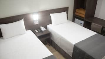 Quarto de hotel com duas camas de solteiro, uma mesa de cabeceira com telefone e luminária, e uma pequena escrivaninha com cadeira.