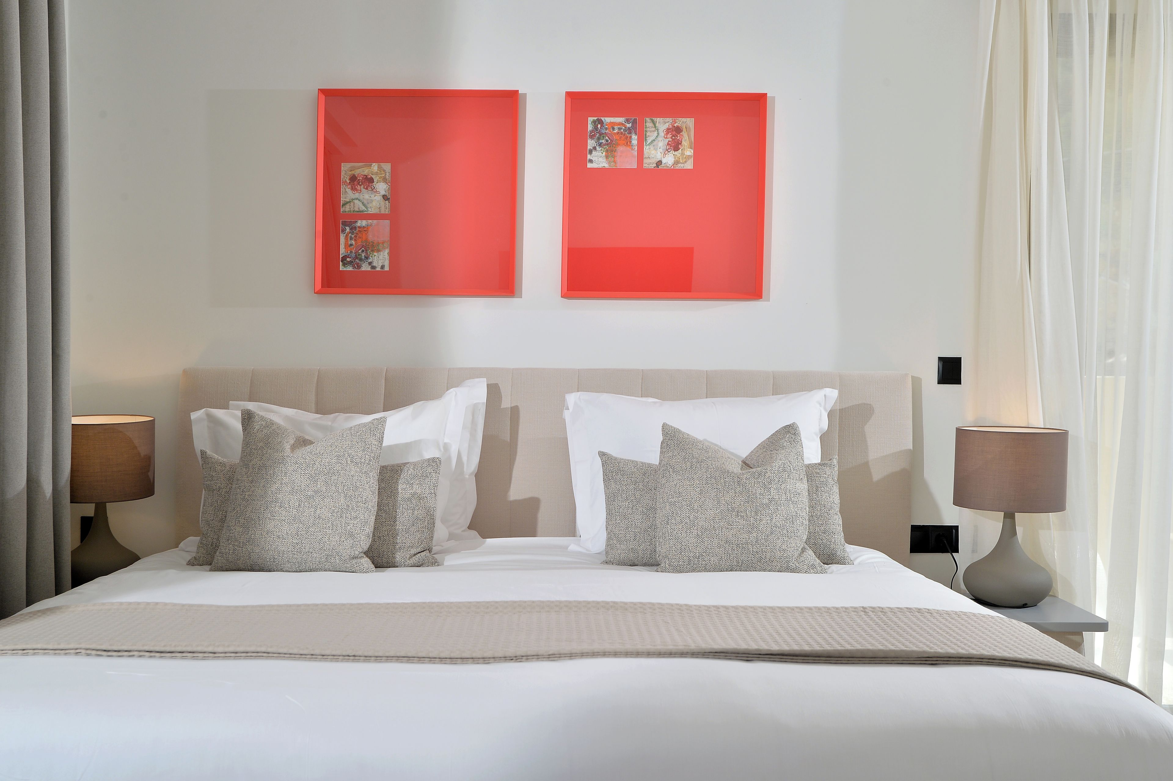 Quarto de hotel com uma cama, duas luminárias de cabeceira, uma janela com cortinas e dois quadros na parede.