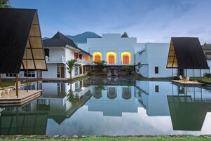 Um hotel com uma piscina refletora, um edifício branco com janelas arqueadas e uma montanha ao fundo.