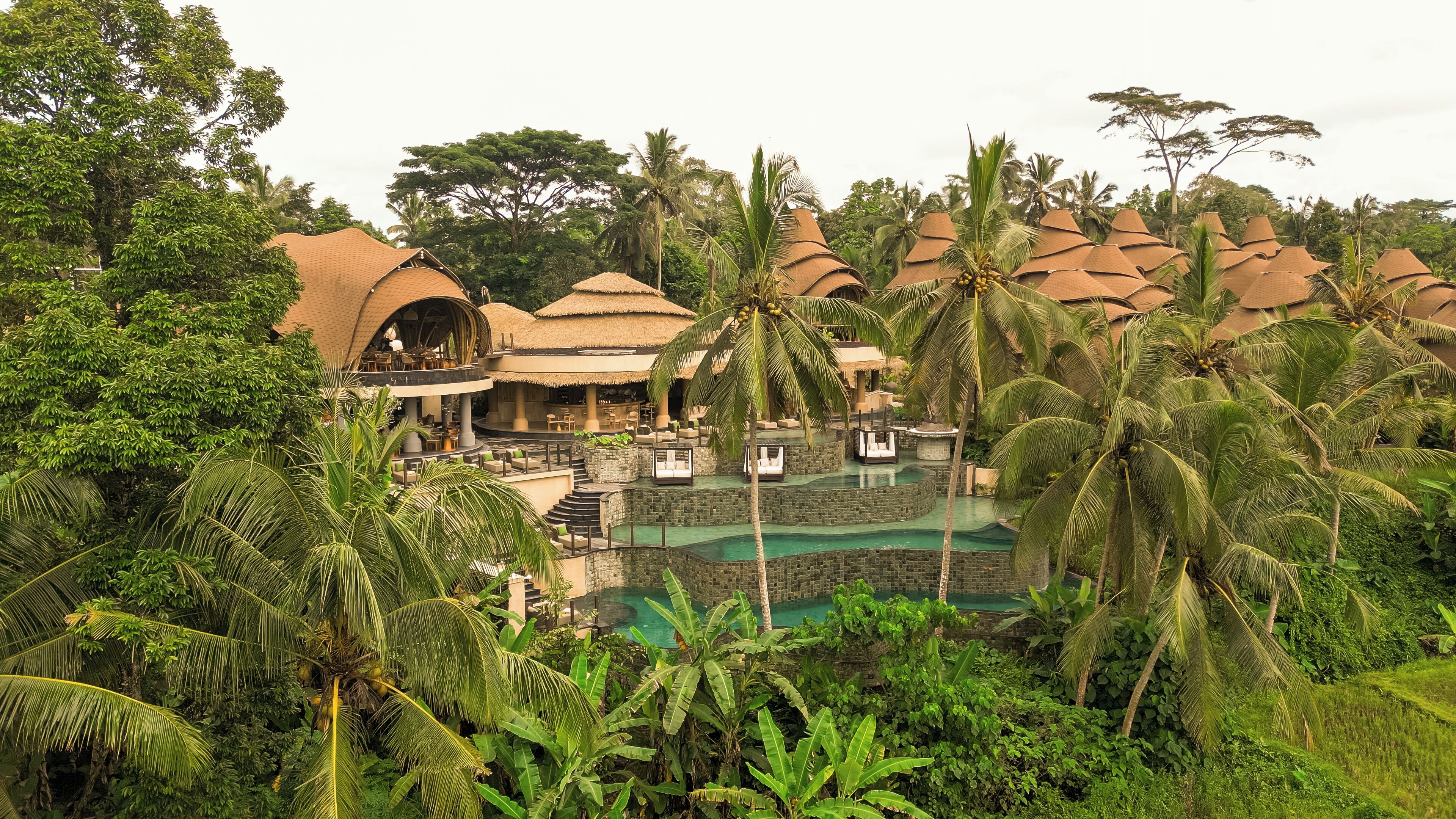 Resort com várias estruturas de telhado de palha, piscina central e vegetação tropical exuberante.