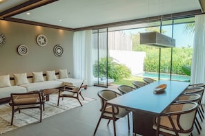 Uma sala de estar moderna com sofá, mesa de jantar e cadeiras, uma grande janela com vista para uma piscina e vegetação.