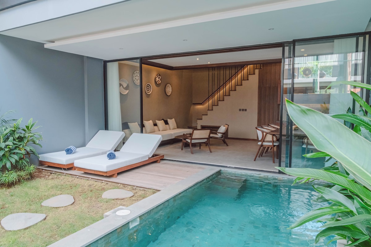 Uma área moderna com piscina ao ar livre, espreguiçadeiras, escada para um andar superior e um espaço de refeições com cadeiras e mesa.
